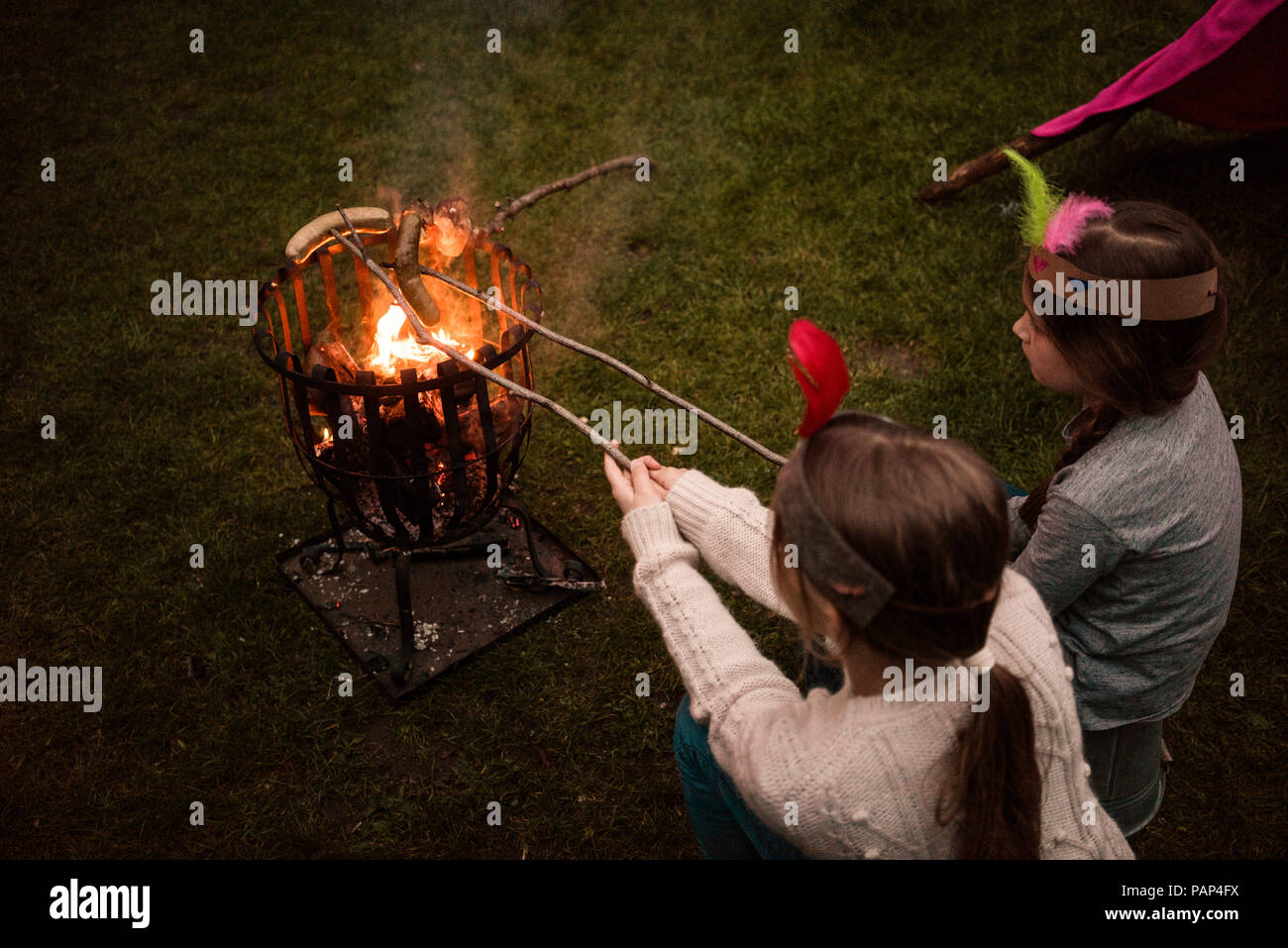 Two girls wearing feather headdress, grilling sausage over camp fire ...