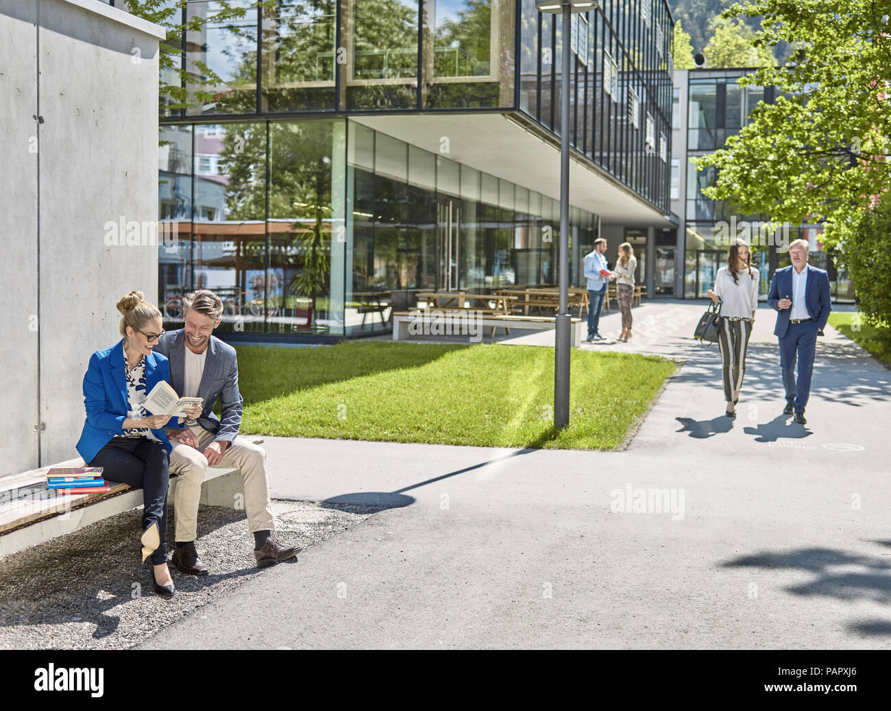 Smiling colleagues with book sitting on bench outside office building ...