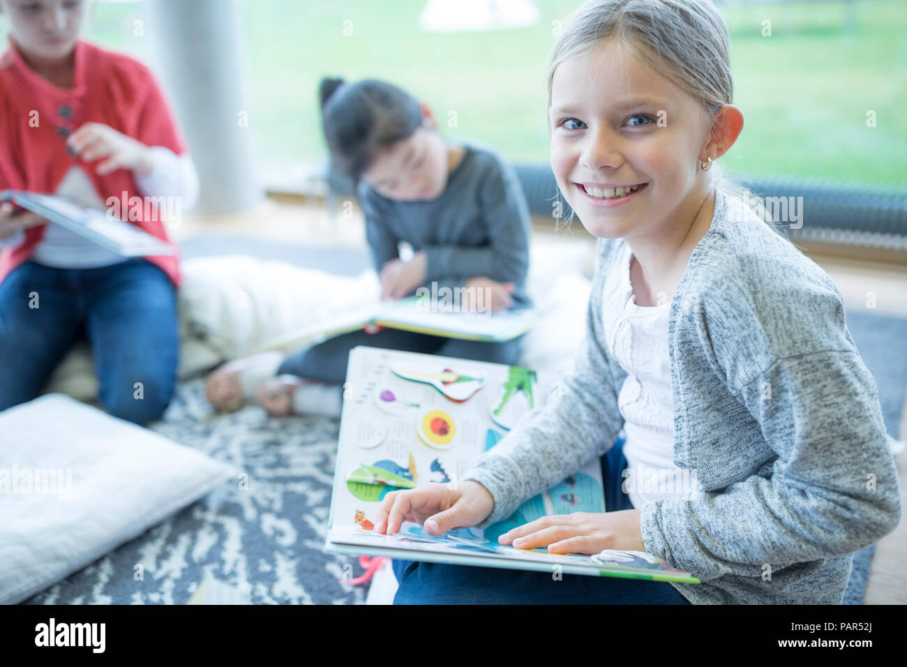 Portrait of smiling schoolgirl sitting on the floor with book in school ...