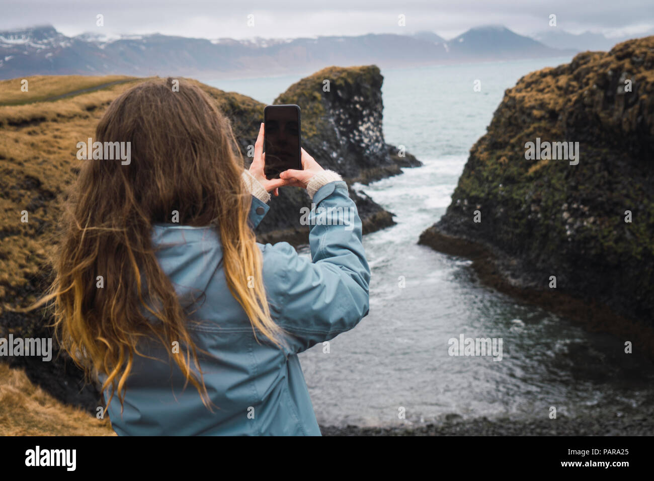 Iceland, back view of young woman taking picture with smartphone at ...