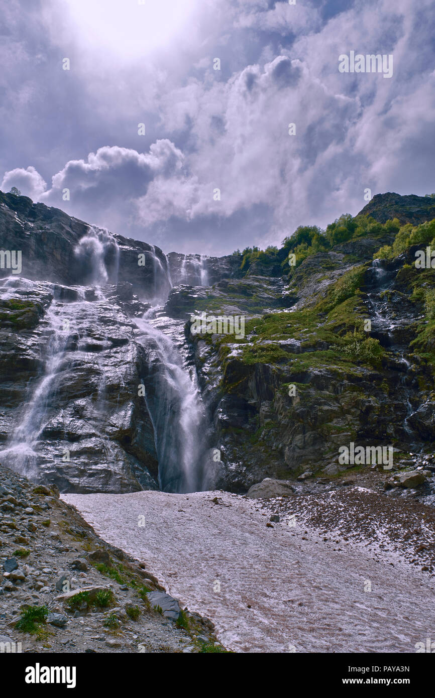 Mountain waterfall under a cloudy sky, lit by sun rays with glacier on ...