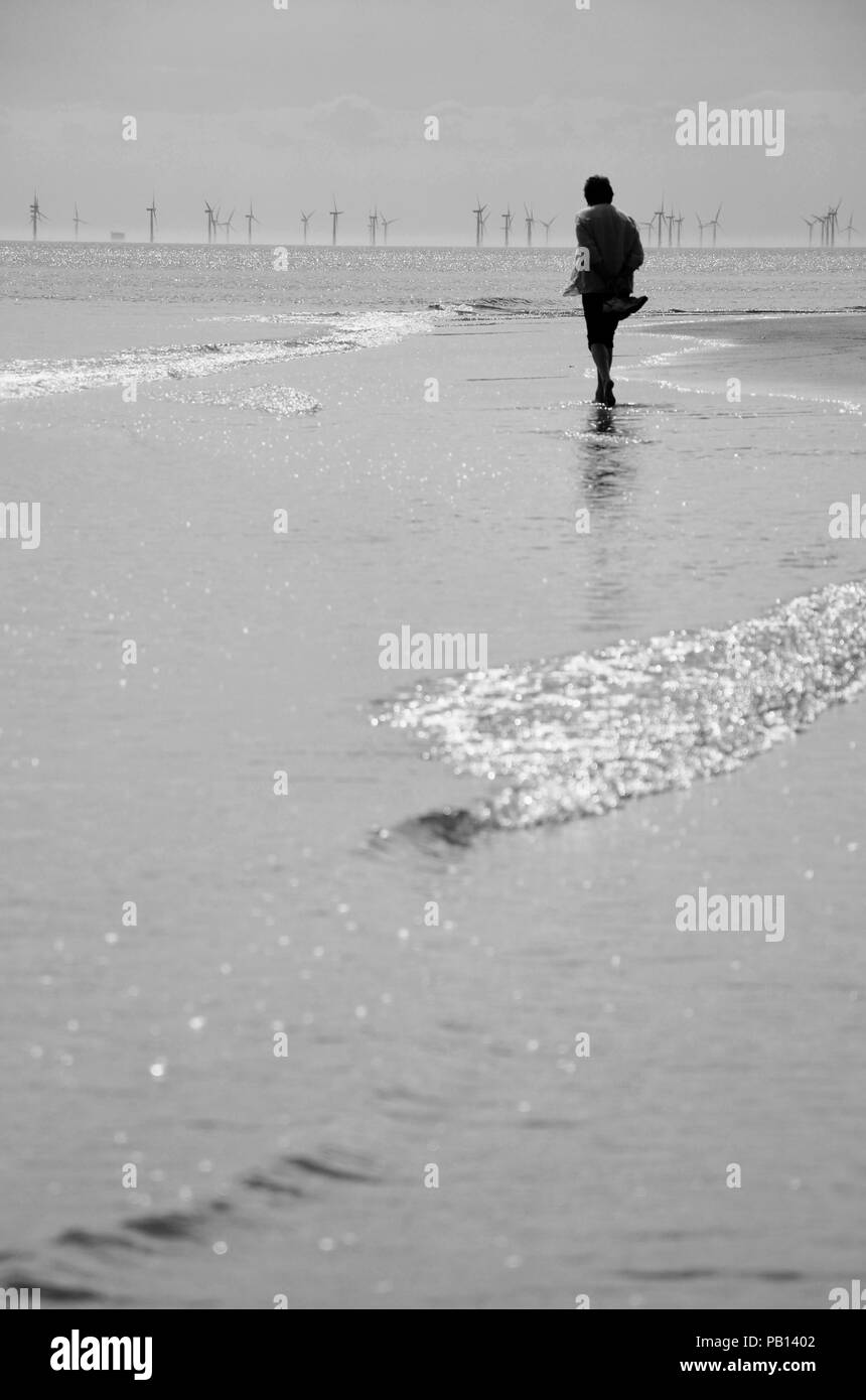 Woman walking alone on the shoreline, wind turbines in distance ...