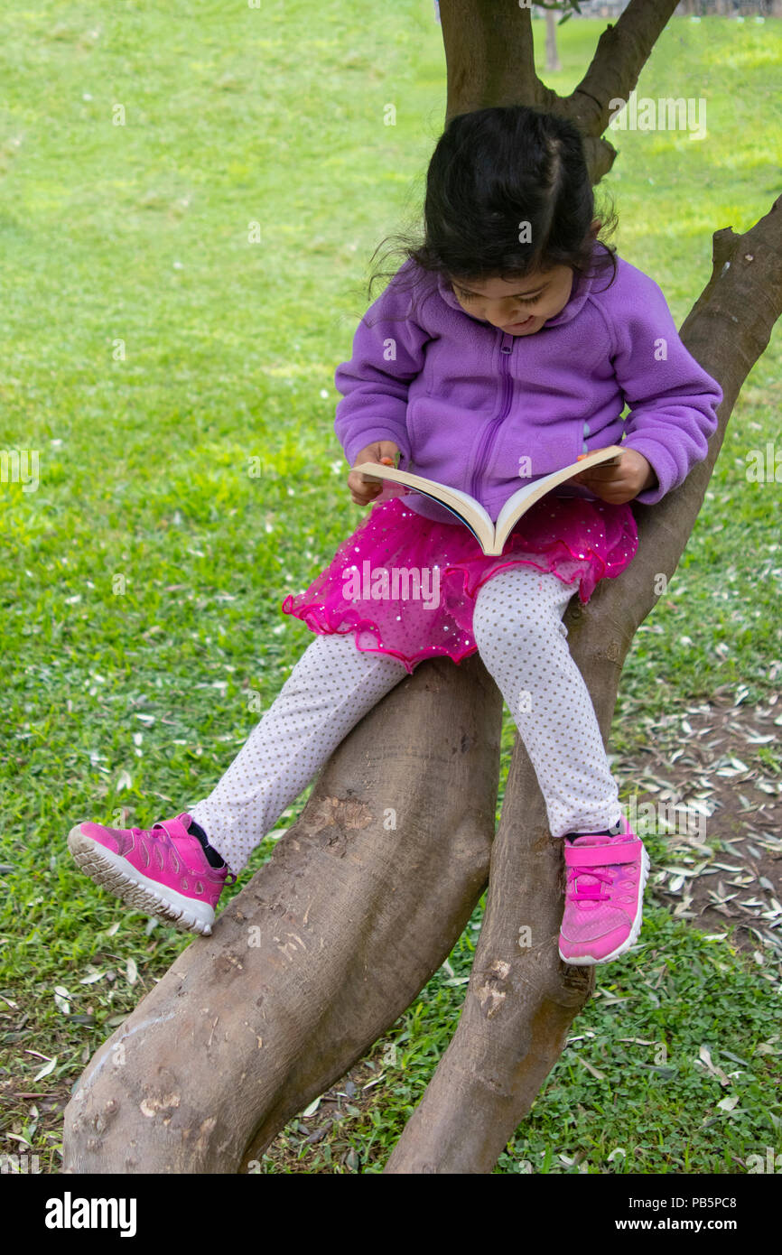 Cute little girl reading a book on a tree in a city park Stock Photo ...
