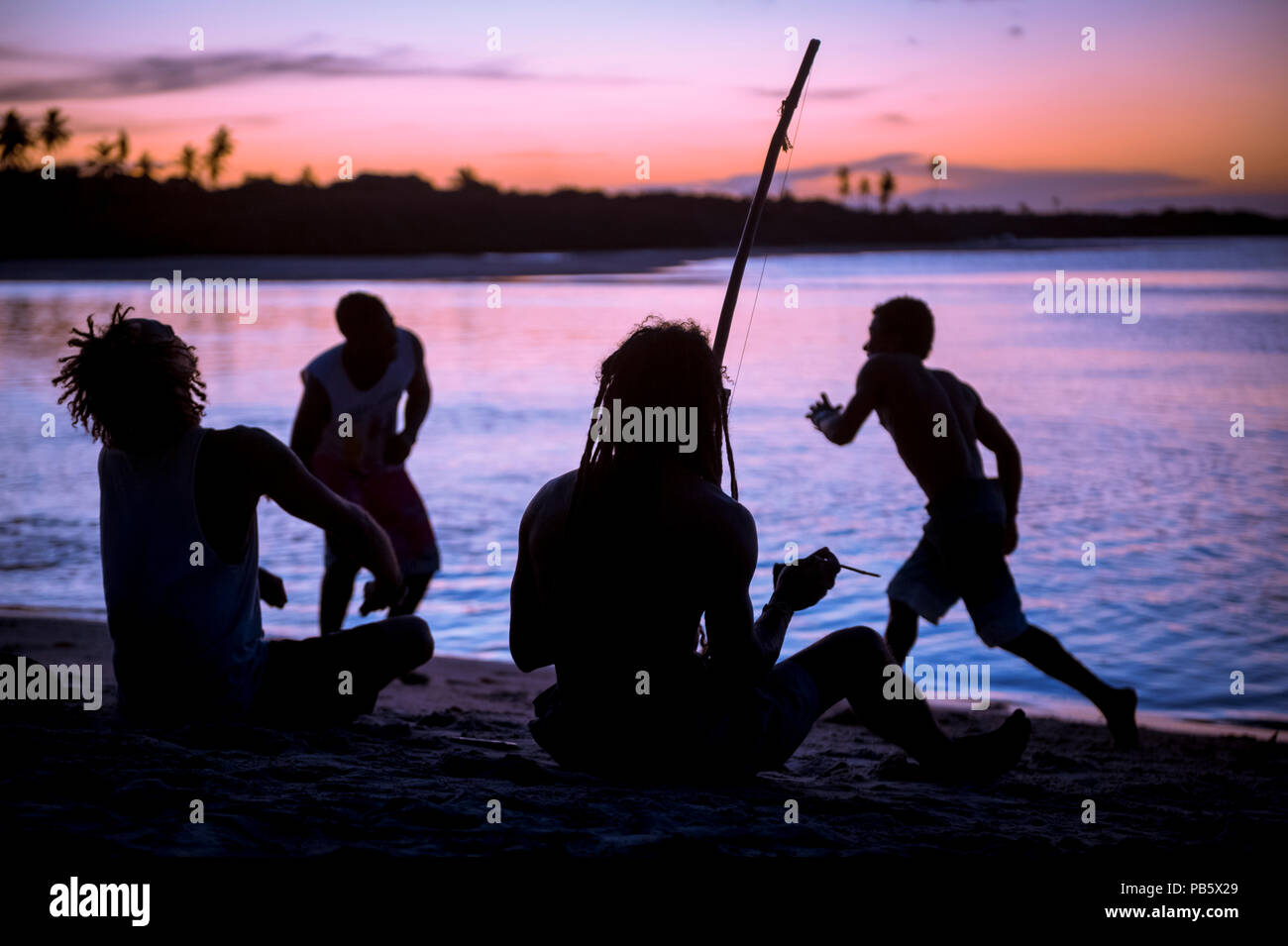Silhouettes of four unrecognizable capoeiristas sat on the beach paying ...