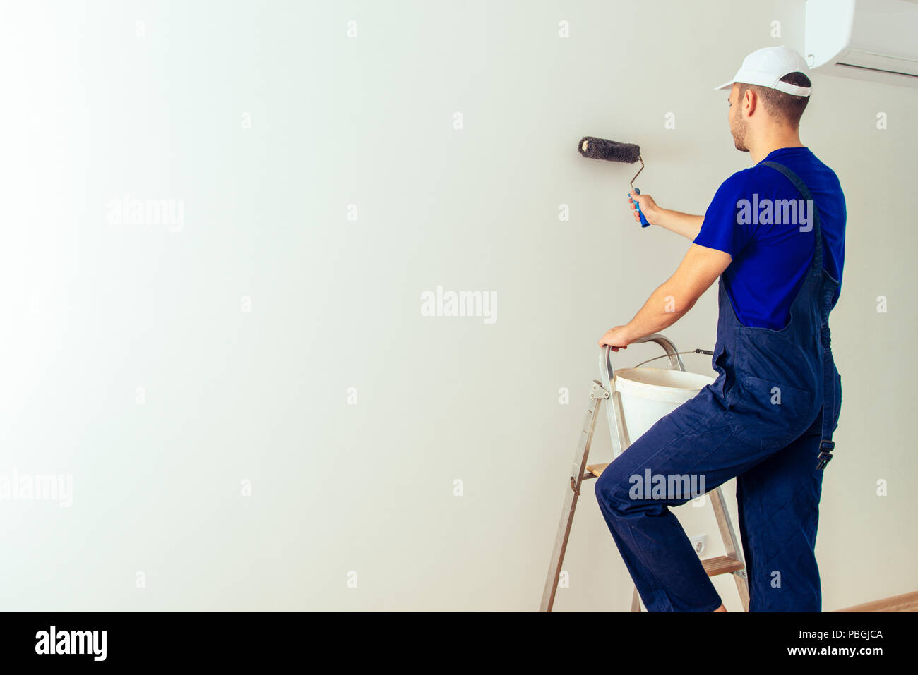 Rear view of painter man looking at white wall, with paint roller and ...
