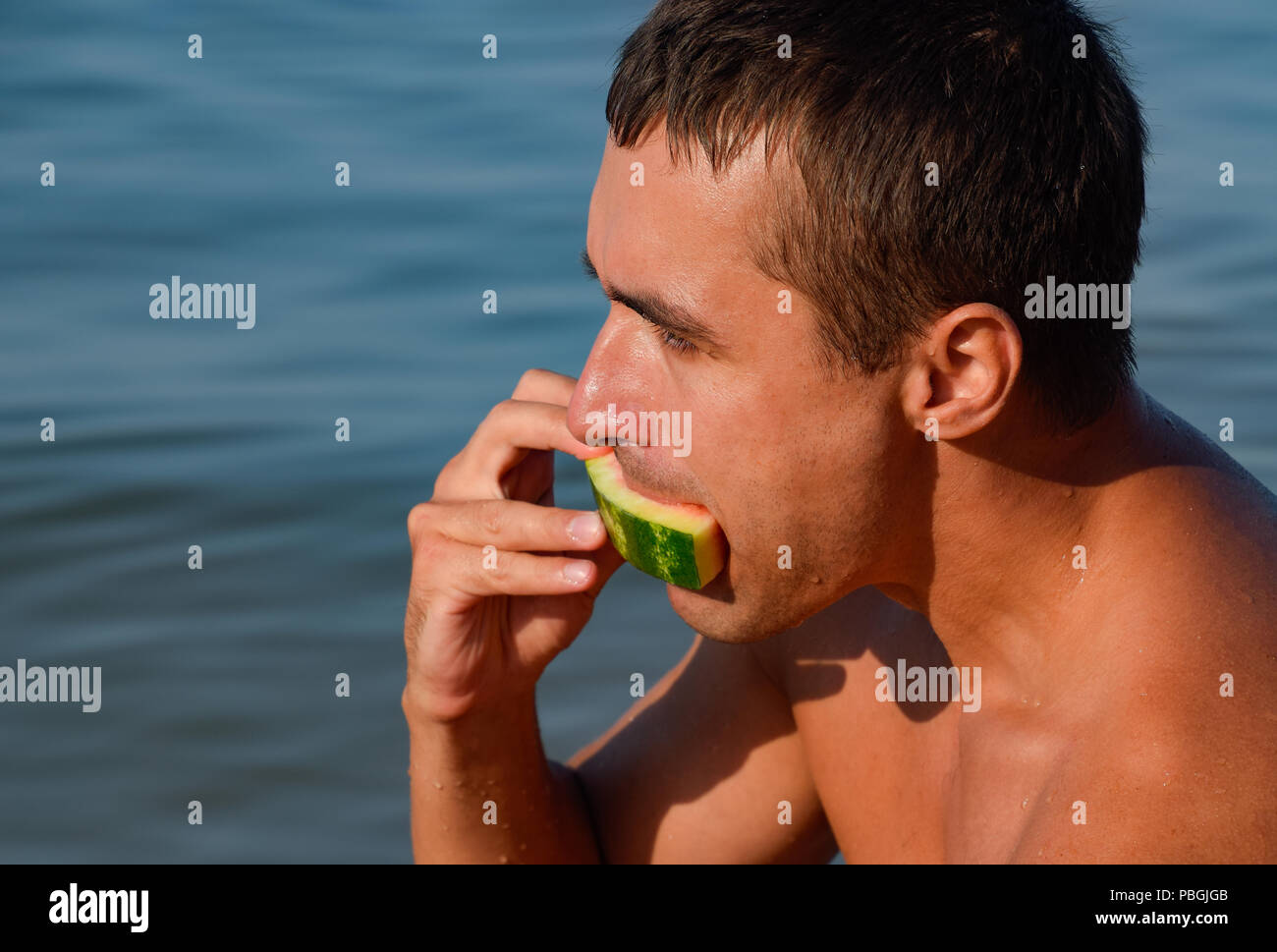 A man is eating a watermelon sitting on the beach. A ripe watermelon in ...