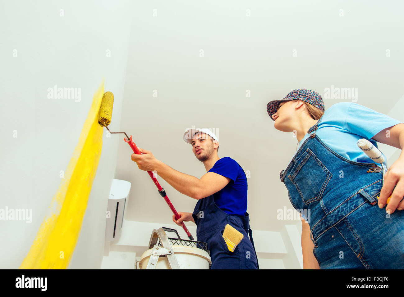 Couple wearing blue overalls painting their living room in yellow color ...