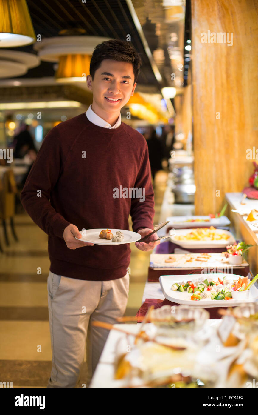 Cheerful young Chinese man taking food from buffet table Stock Photo ...