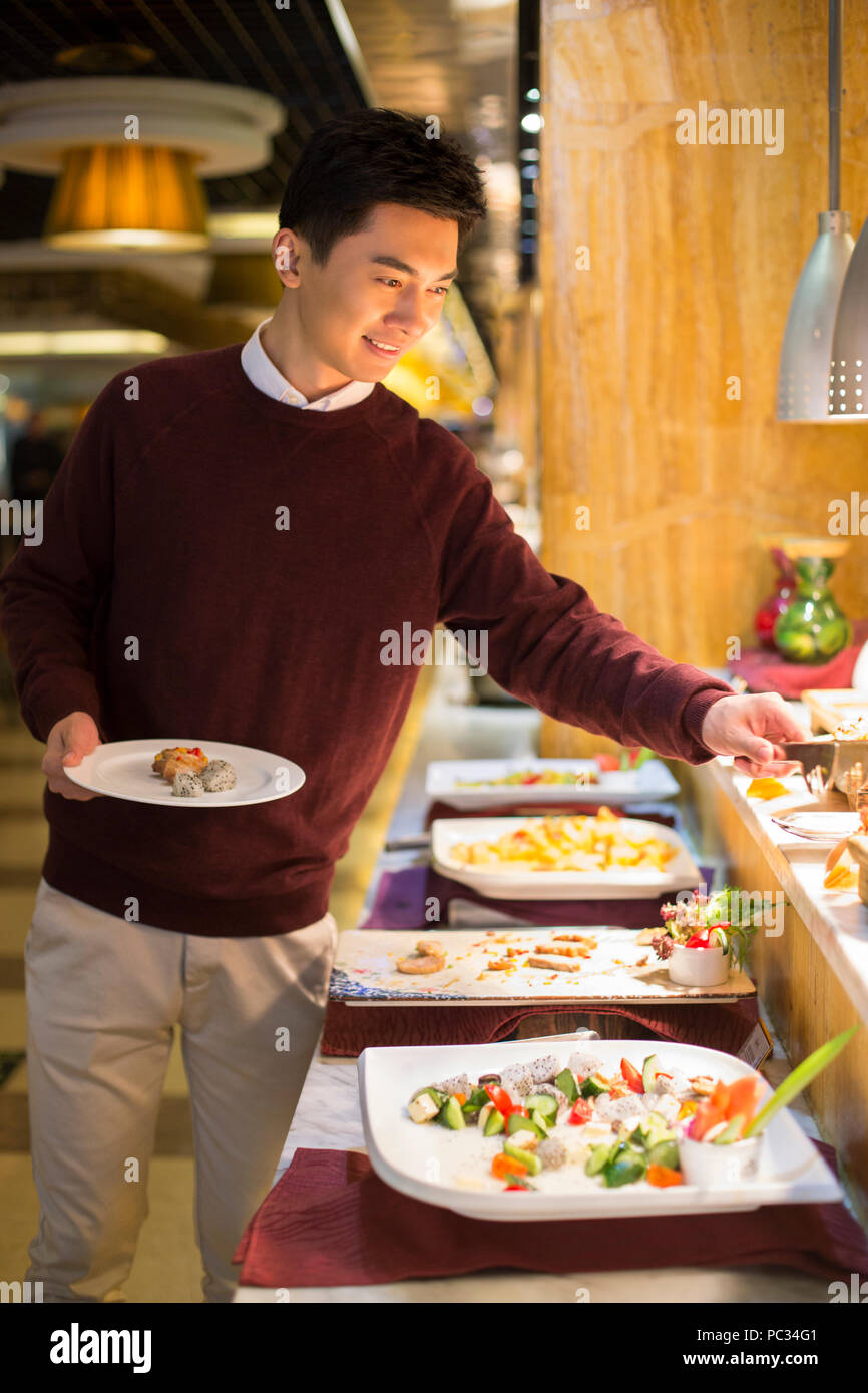 Cheerful young Chinese man taking food from buffet table Stock Photo ...