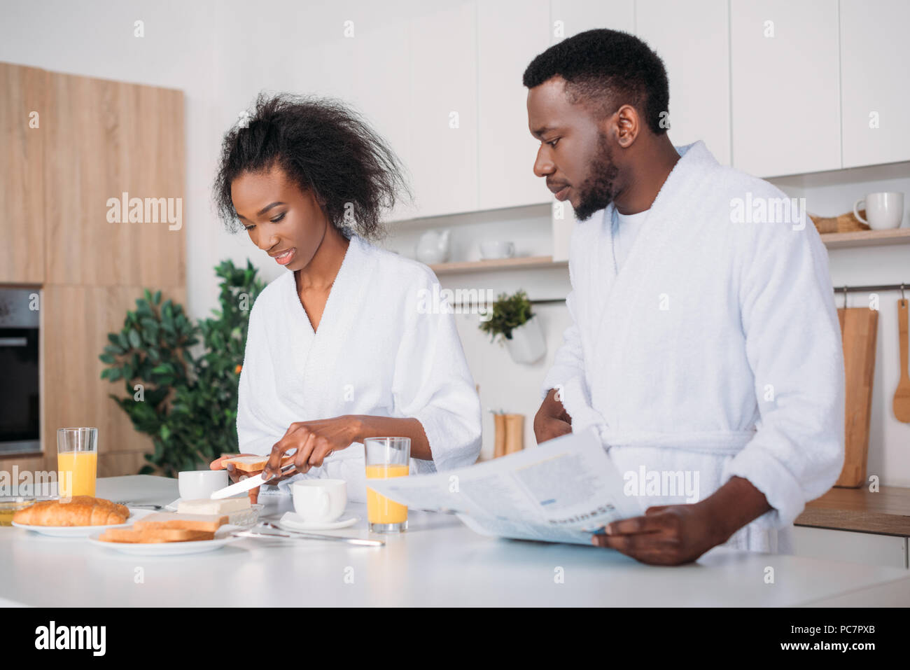 Young woman spreading butter on toast while man standing with newspaper ...