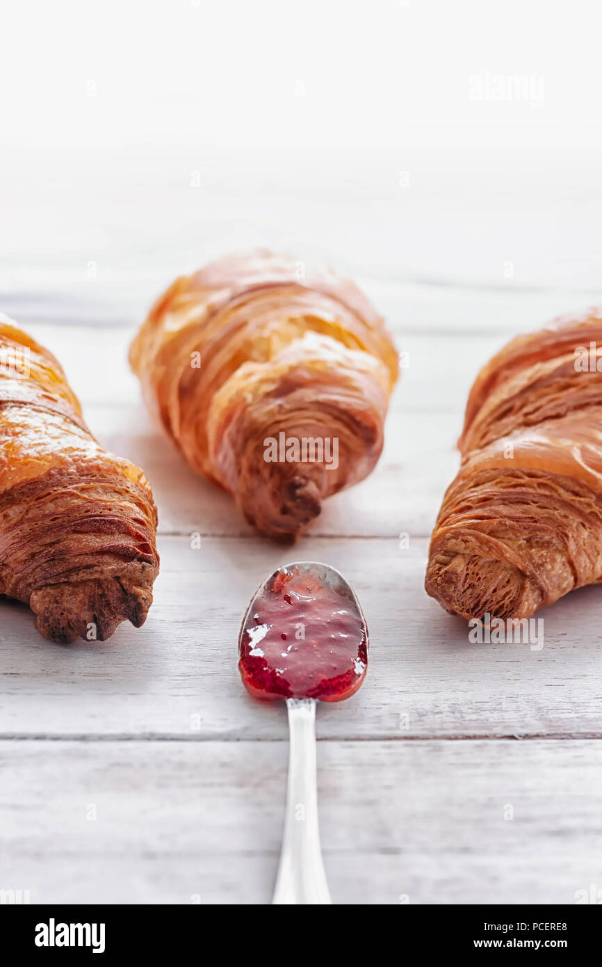 Three croissants on a white wooden table with spoon filled with jam in ...