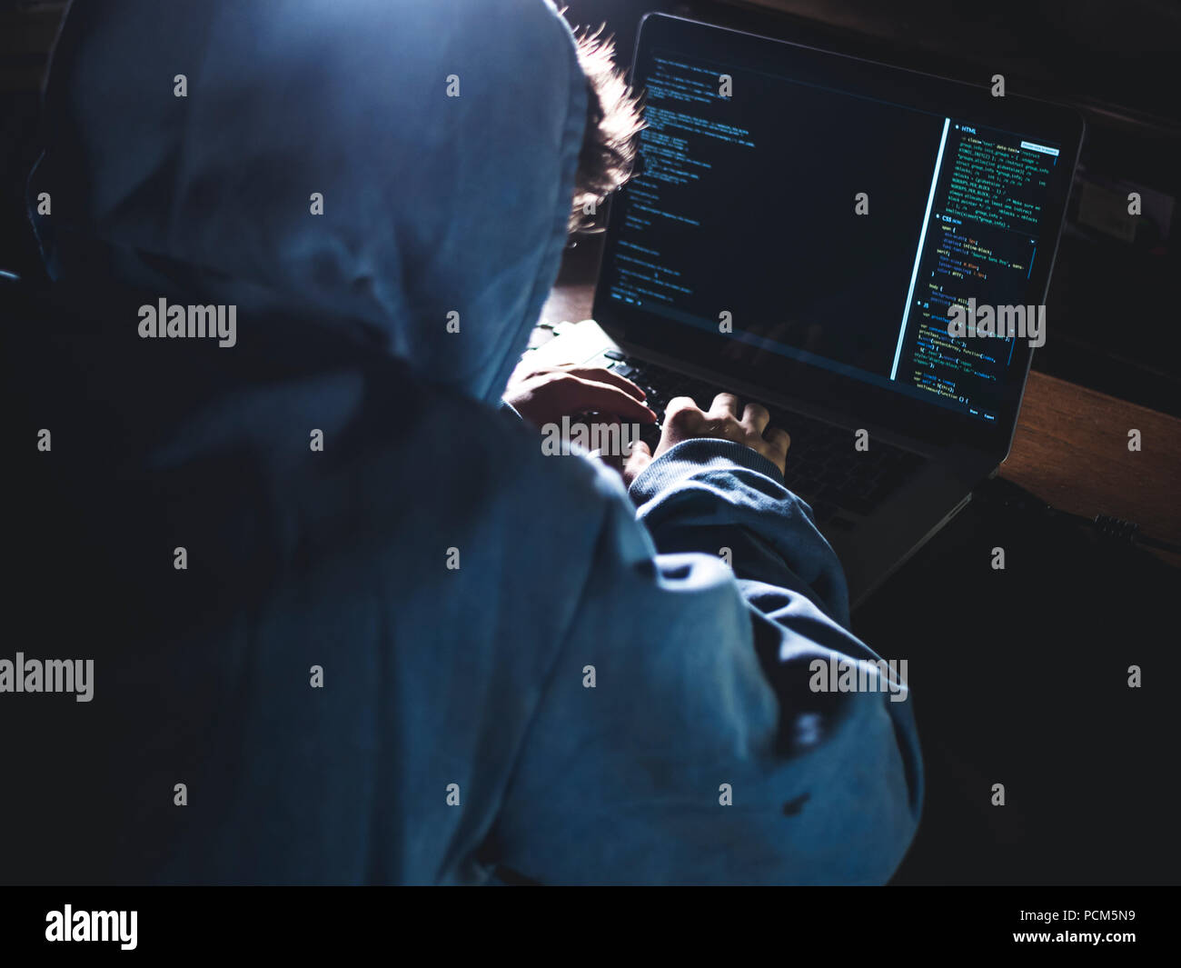 hacker in hoodie typing code in program using laptop in dark studio ...