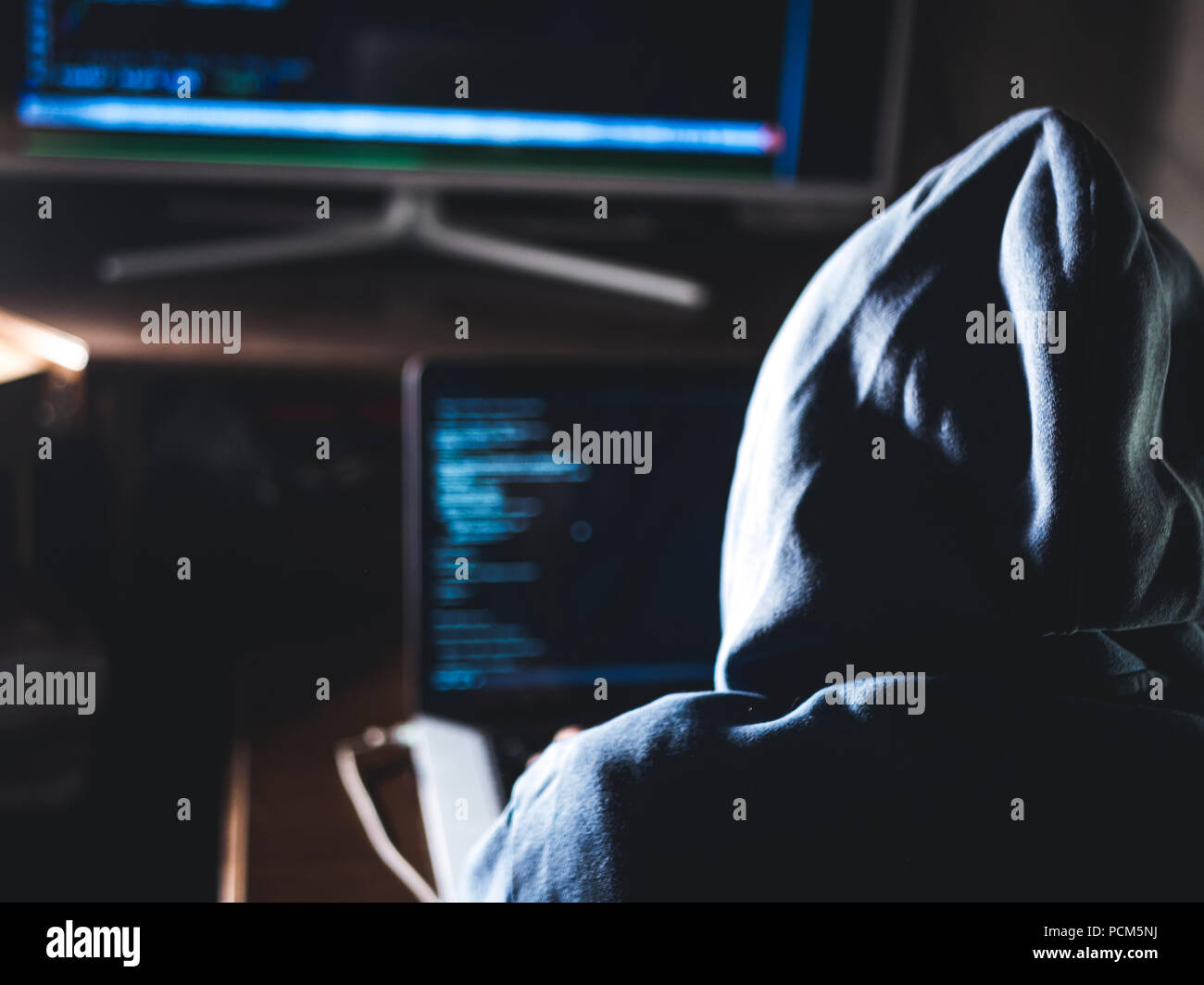 hacker in hoodie typing code in program using laptop in dark studio ...
