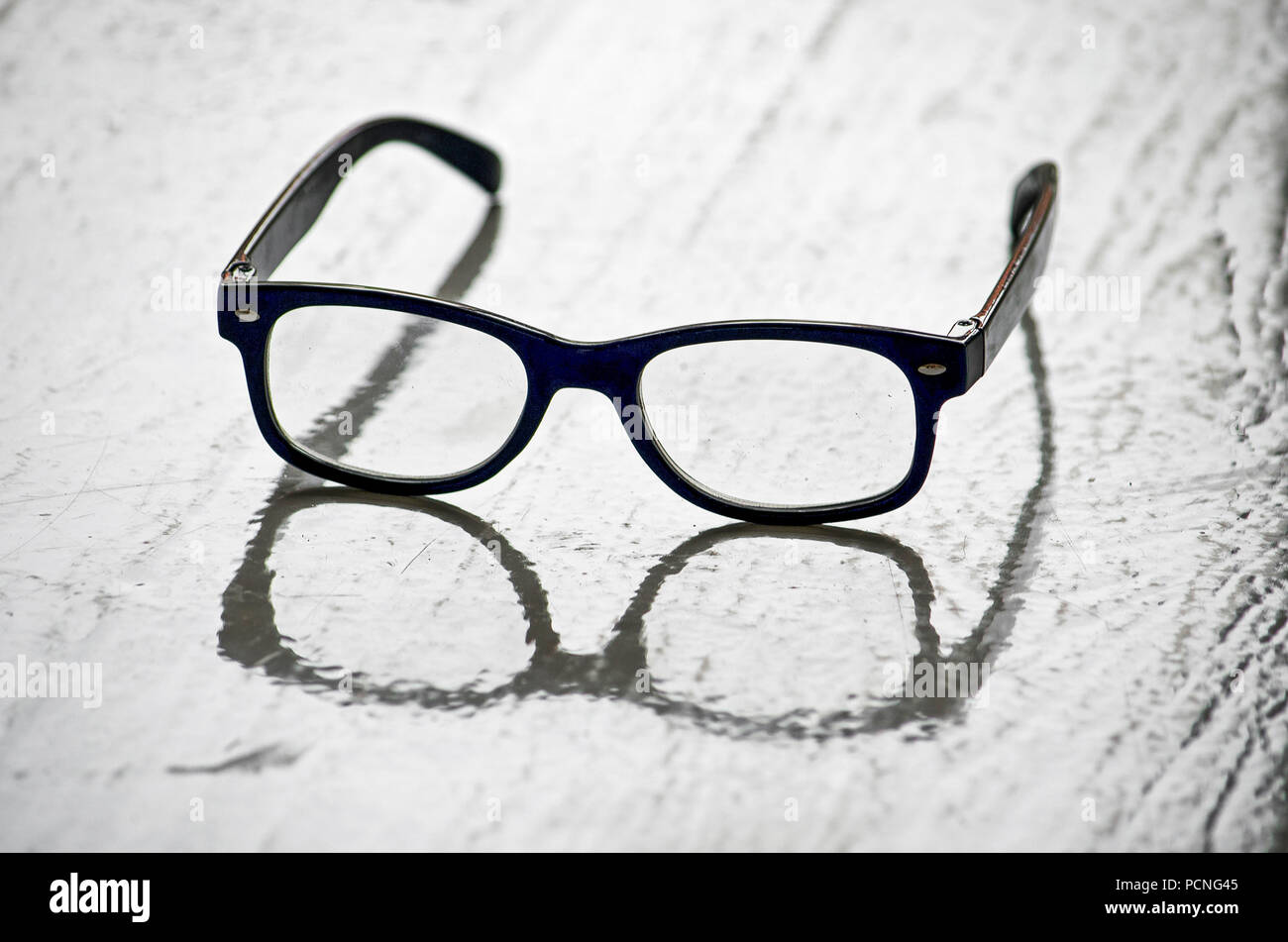 A black pair of simple reading glasses on a reflective white table ...
