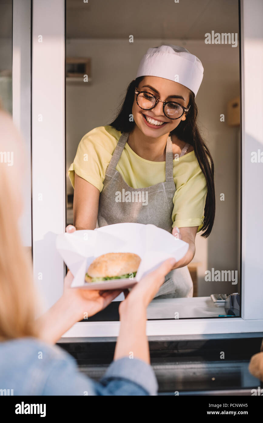 cropped image of smiling chef giving burger to customer in food truck ...