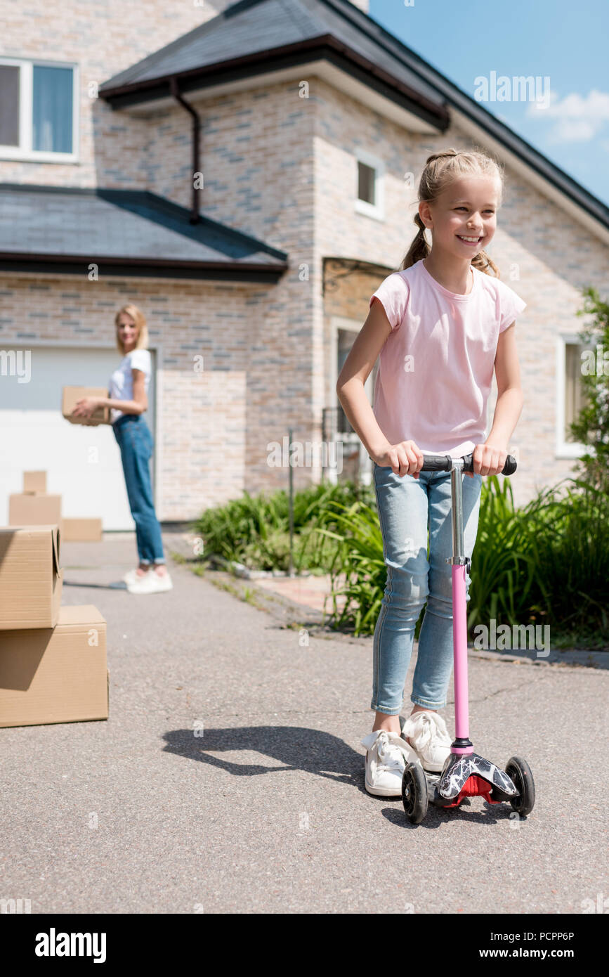 smiling little child riding on kick scooter while her mother unpacking ...