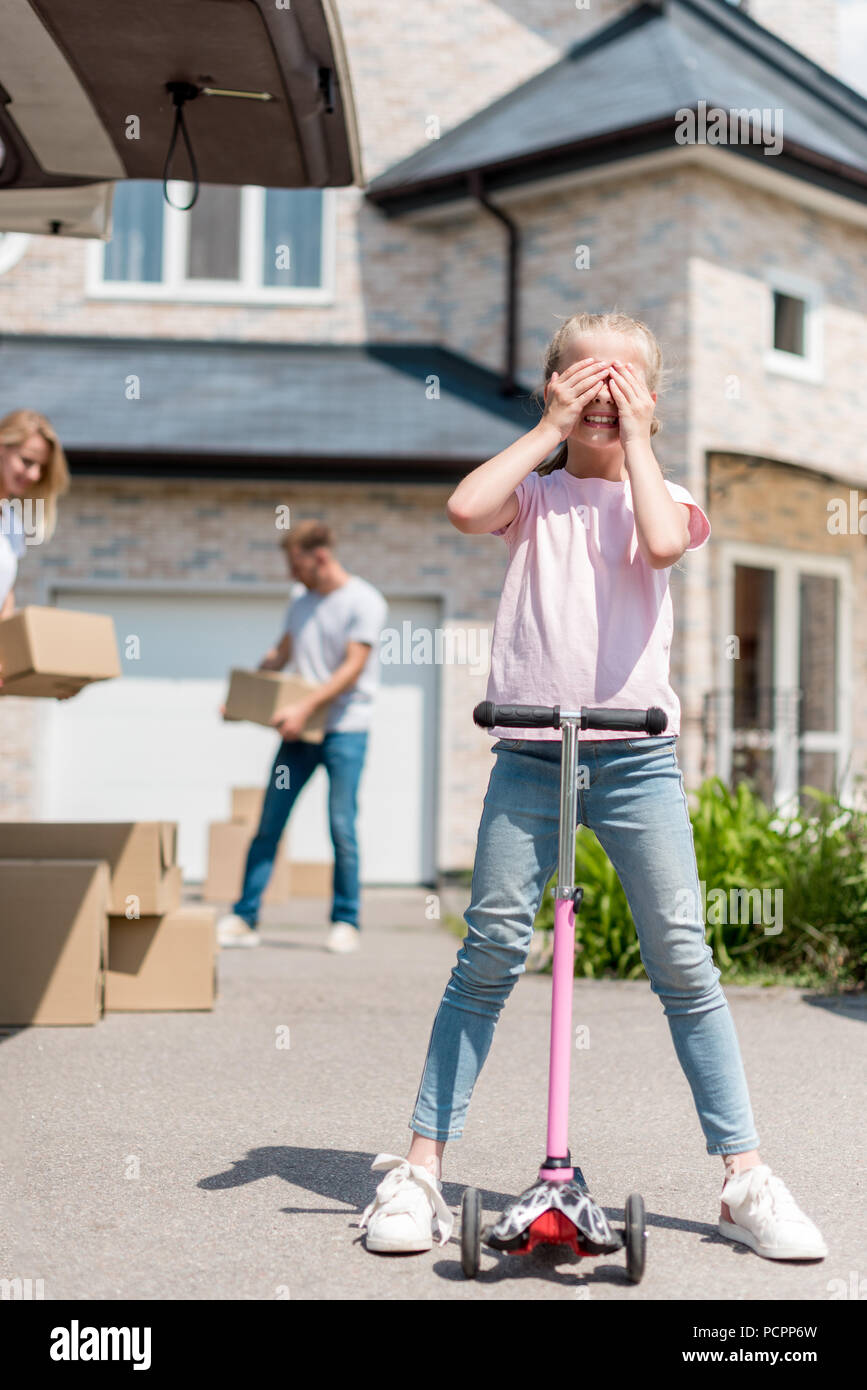 little kid covering eyes on kick scooter and her parents unpacking ...