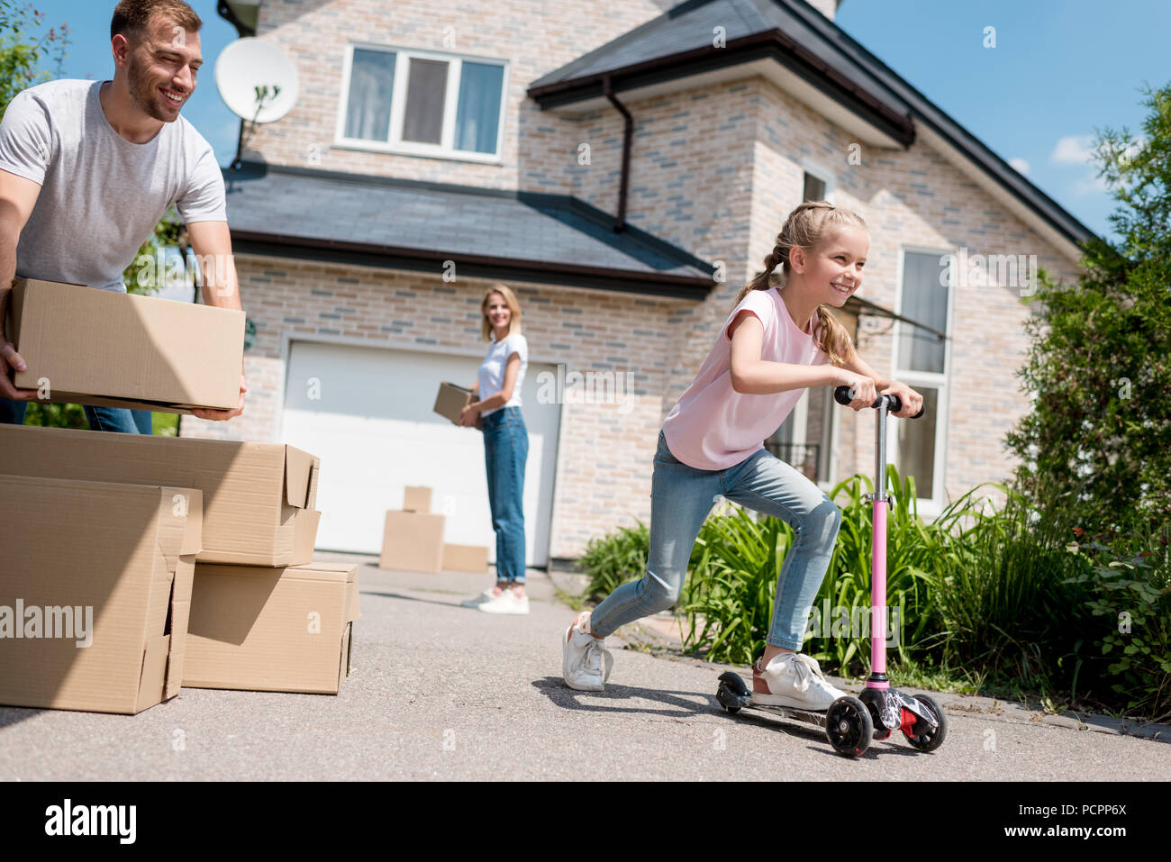 little kid riding on kick scooter and her parents unpacking cardboard ...