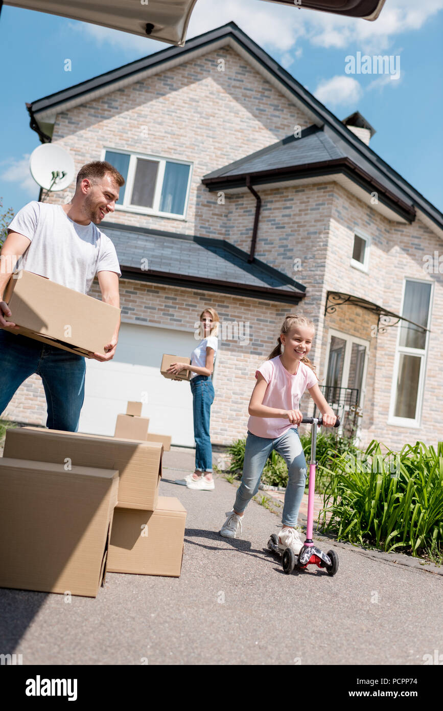 smiling little kid riding on kick scooter and her parents unpacking ...