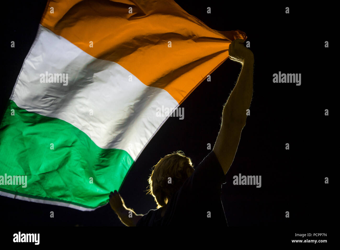 Man waving Irish tricolor flag at night backlit by bright floodlights ...