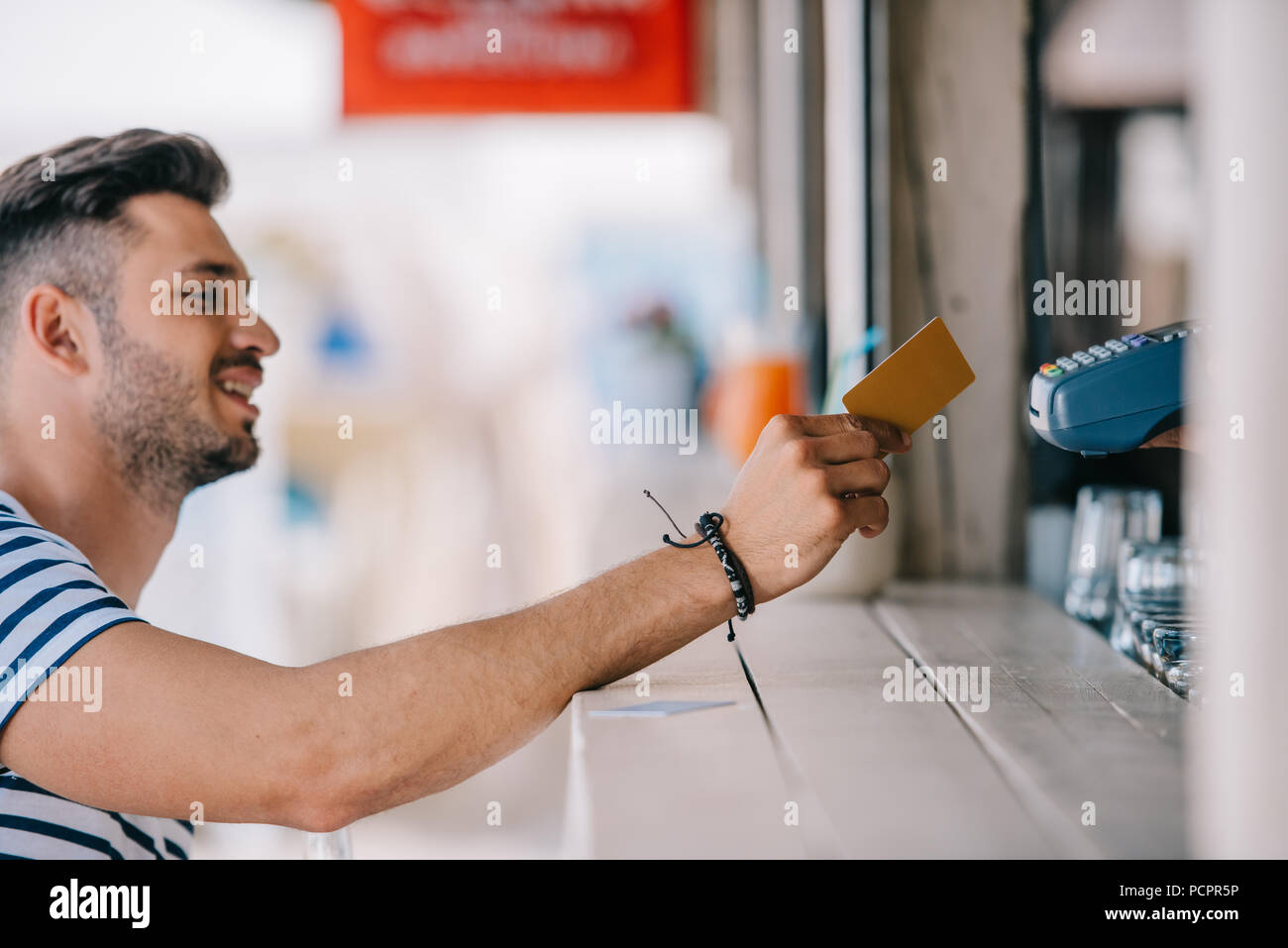 side view of smiling young man paying with credit card at beach bar ...