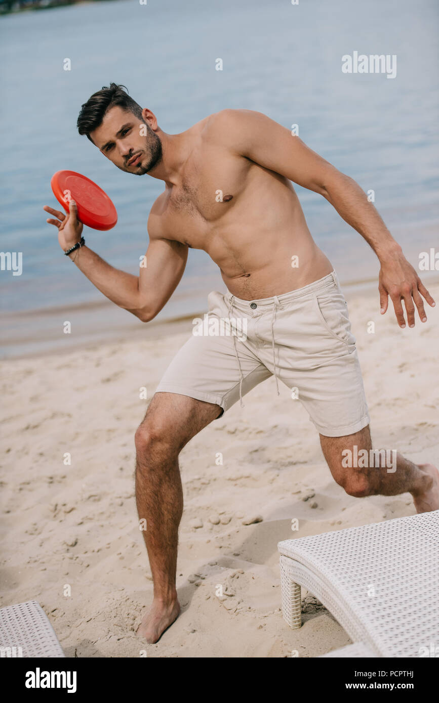 handsome young man holding flying disk and looking at camera on sandy ...