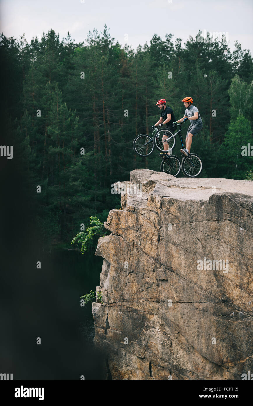 side view of trial bikers standing on back wheels on rocky cliff with ...