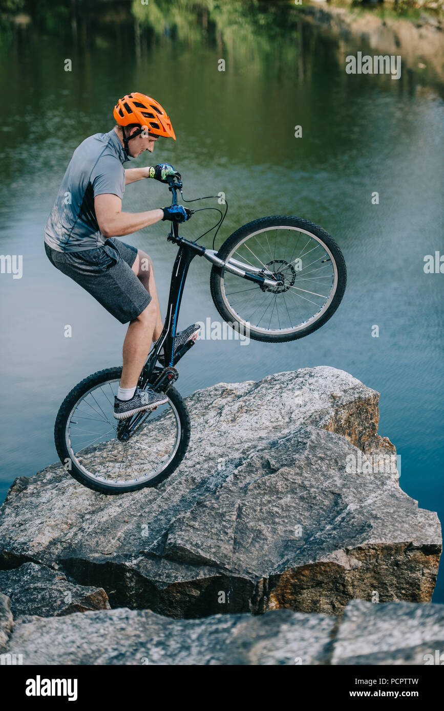 extreme trial biker balancing on back wheel on rocky cliff over lake ...