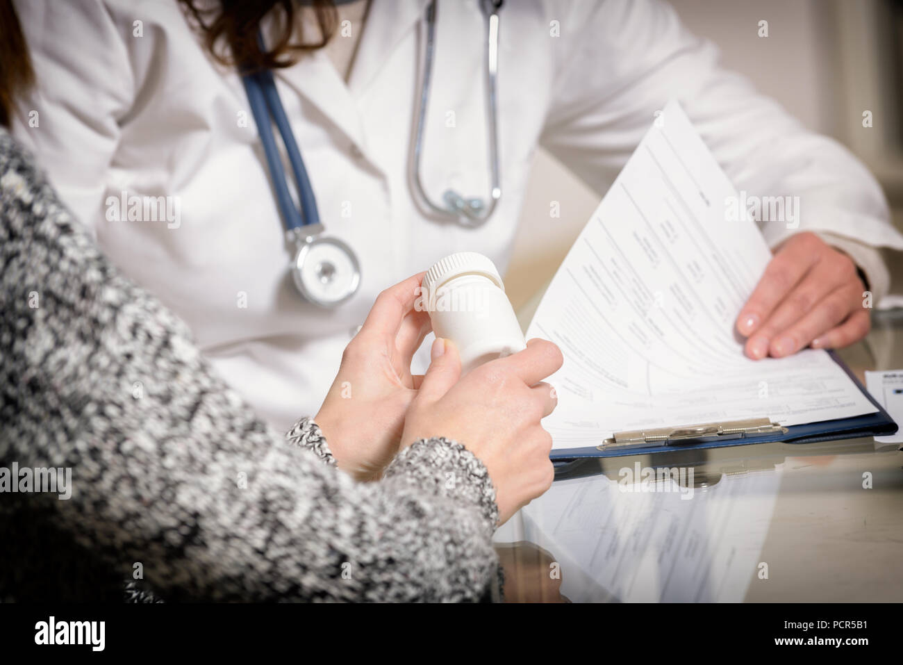 Patient holding a bottle of pills and doctor making notes in background ...