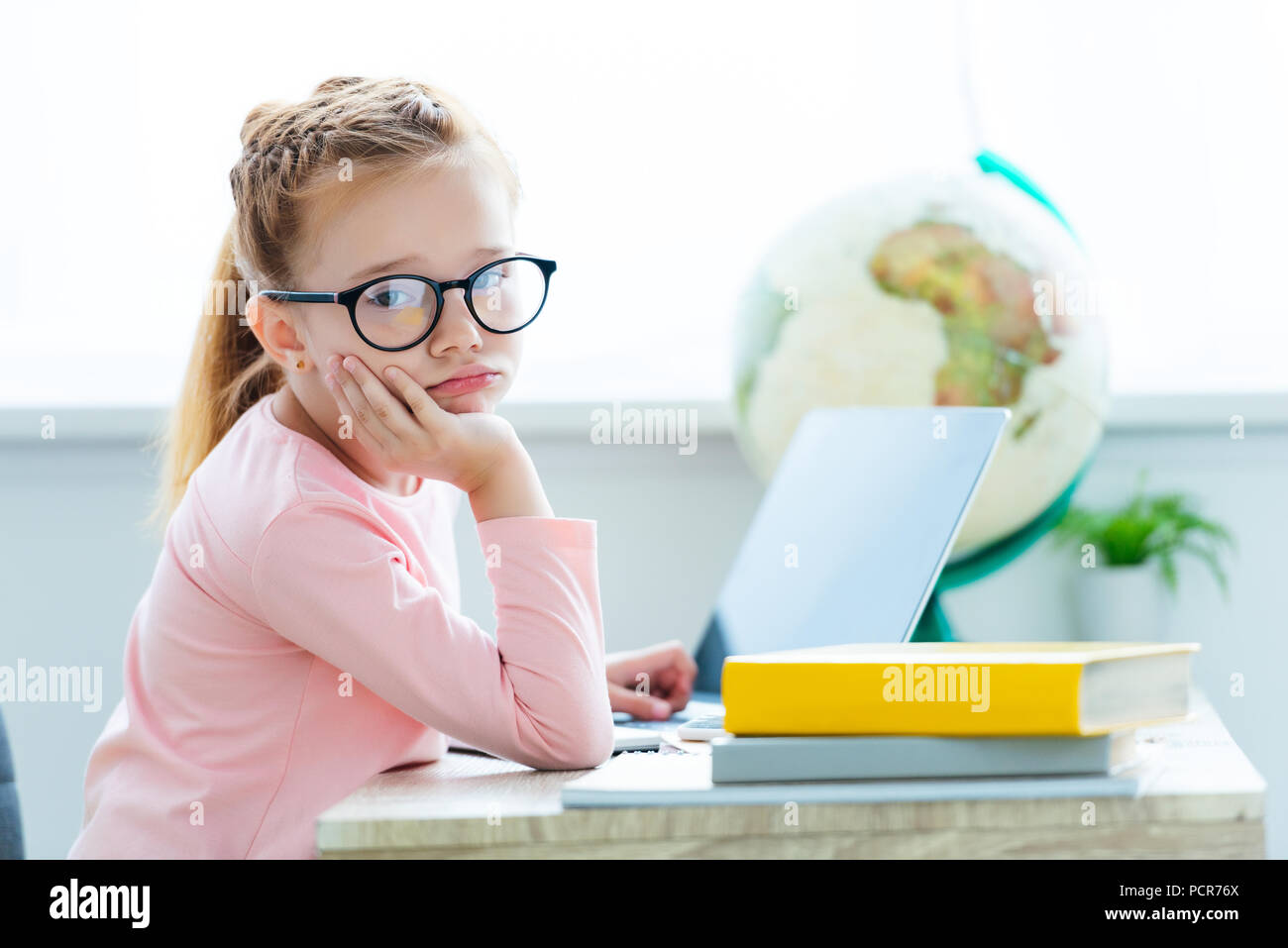 bored child in eyeglasses looking at camera while studying with laptop ...