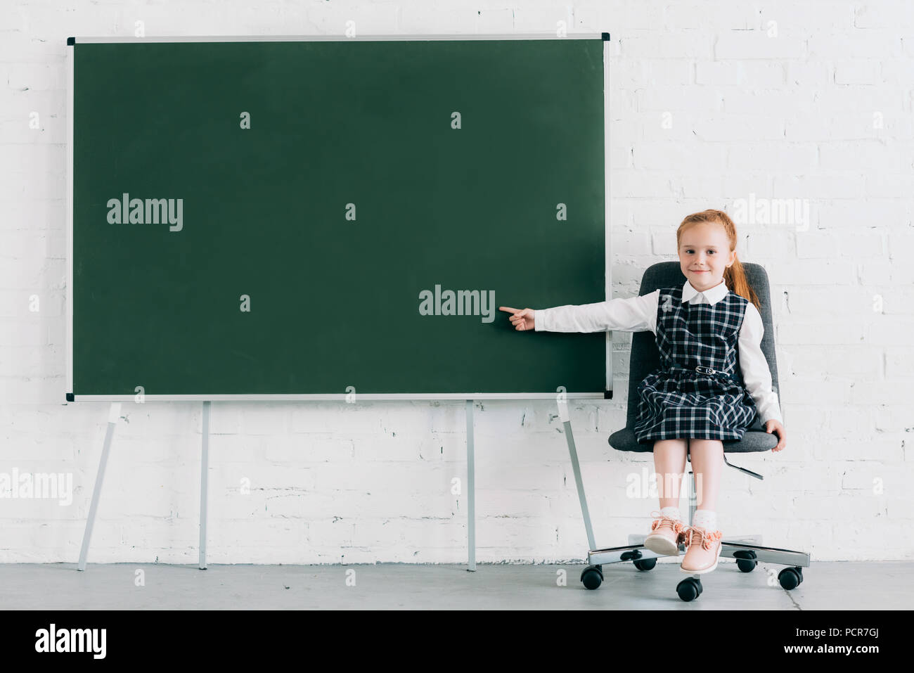 adorable little schoolgirl smiling at camera and pointing at blank chalkboard while sitting on ...