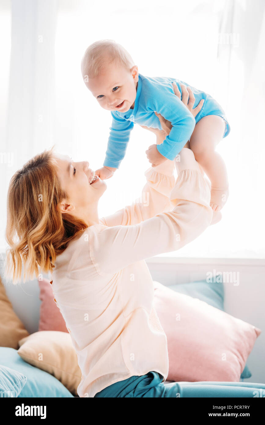 side view of mother raising laughing little child while sitting on bed ...