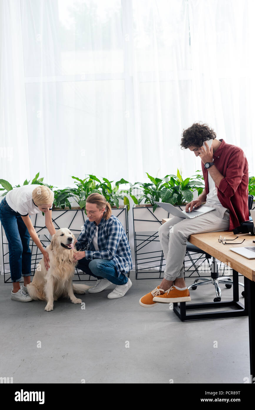 young man talking by smartphone while colleagues stroking dog in office ...