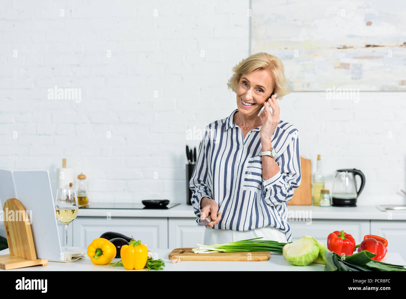 smiling attractive grey hair woman cutting green onion in kitchen and ...
