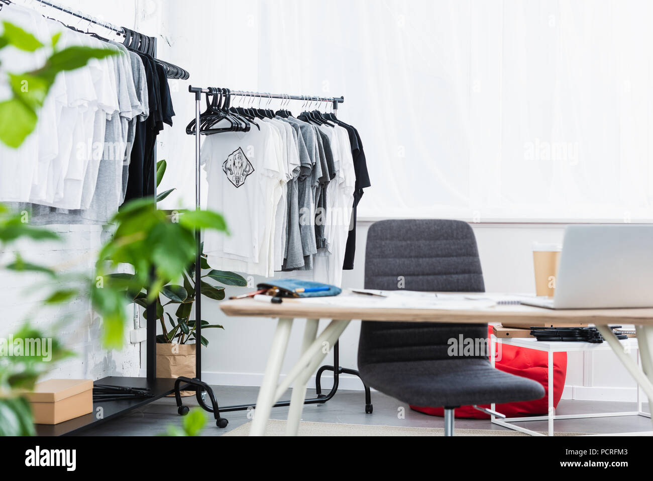 selective focus of t-shirts on hangers and working table with laptop in ...