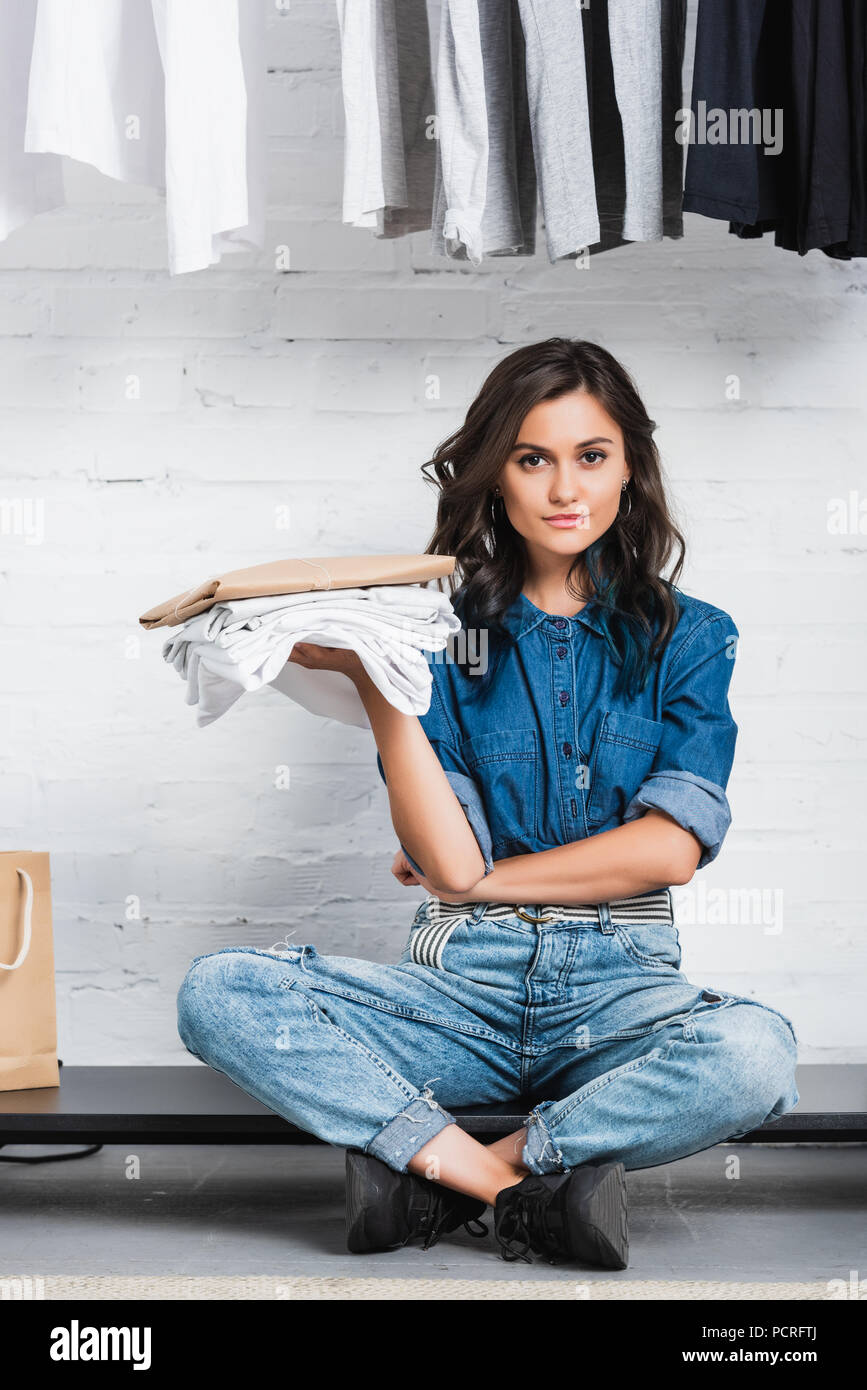 female designer sitting with stack of white t-shirts and paper package ...