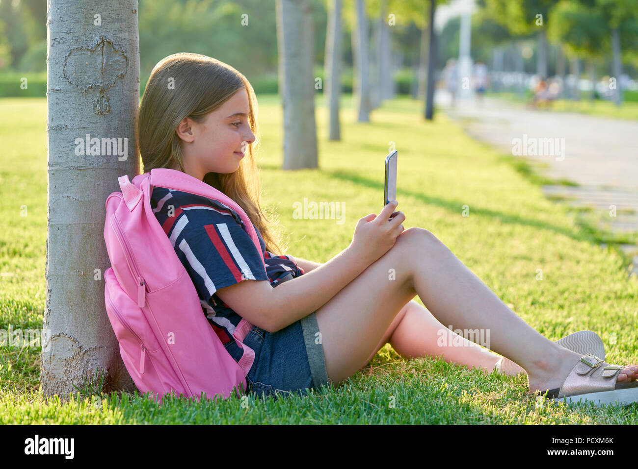 Blond student kid girl with smartphone in a park back to school sit on ...