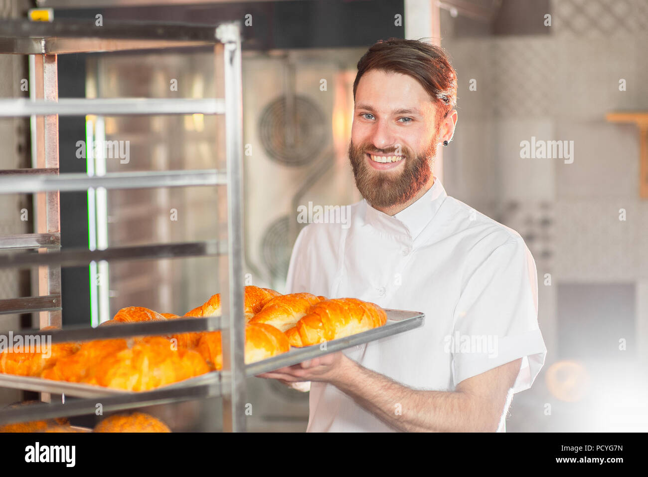 A young handsome baker takes out hot fresh baking from the oven in the ...