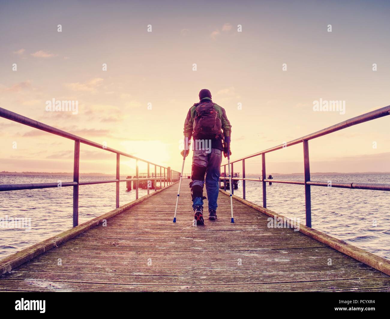 Hurt man with hooded jacket and forearm crutches standing on sea bridge ...