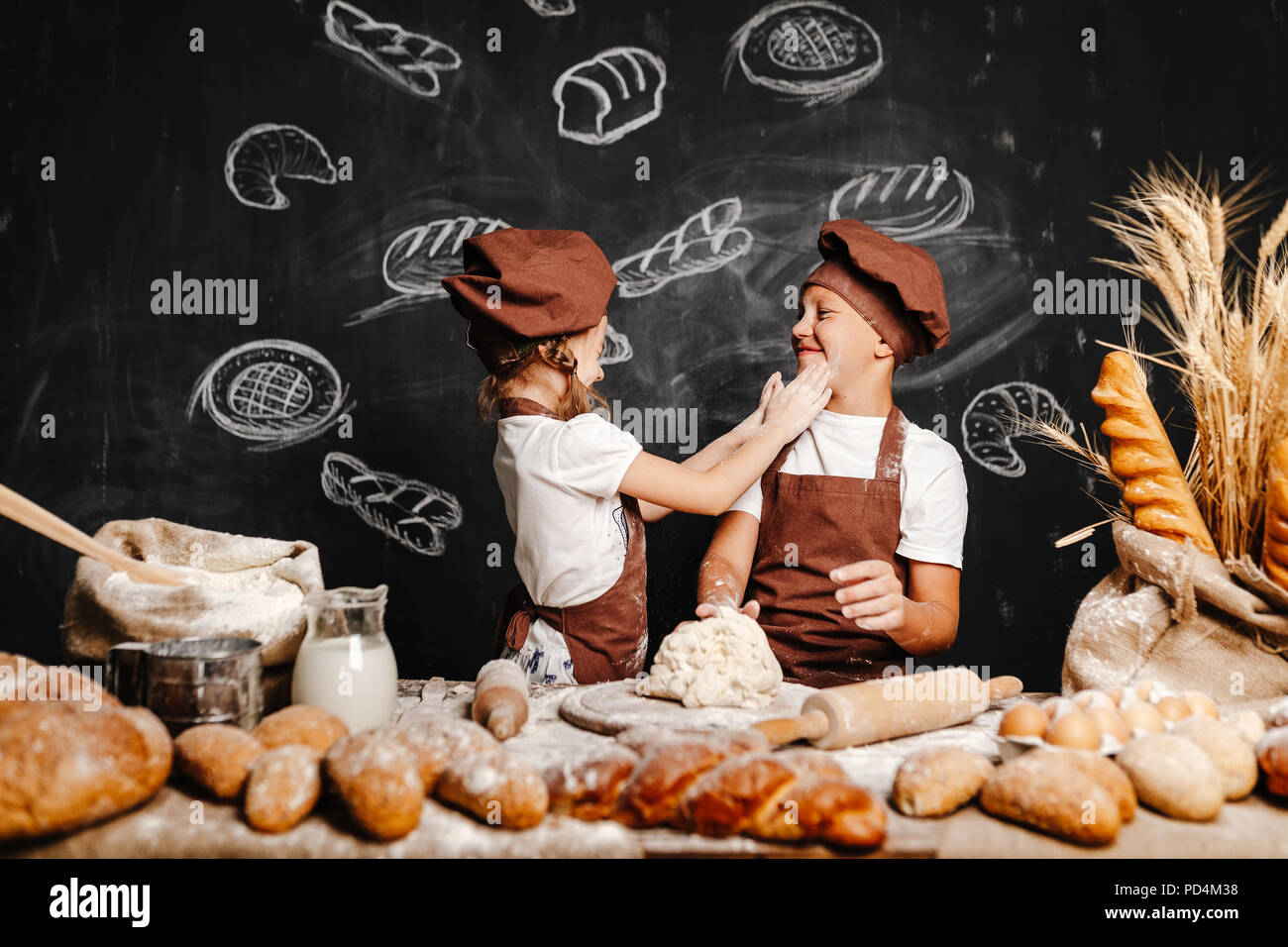 Adorable girl with brother in aprons on table with bread loaves making ...