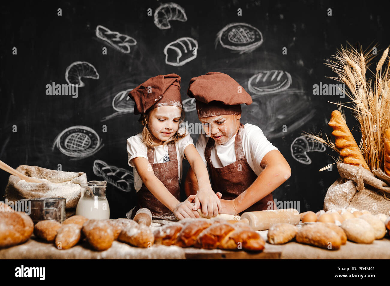 Adorable girl with brother in aprons on table with bread loaves making ...