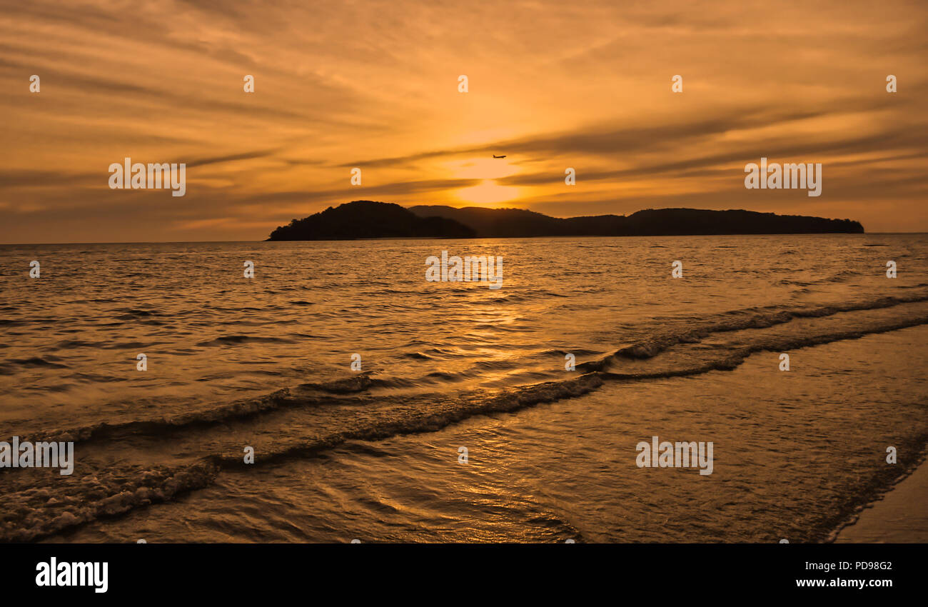 A plane flies directly over an island by the sea in the light of the ...