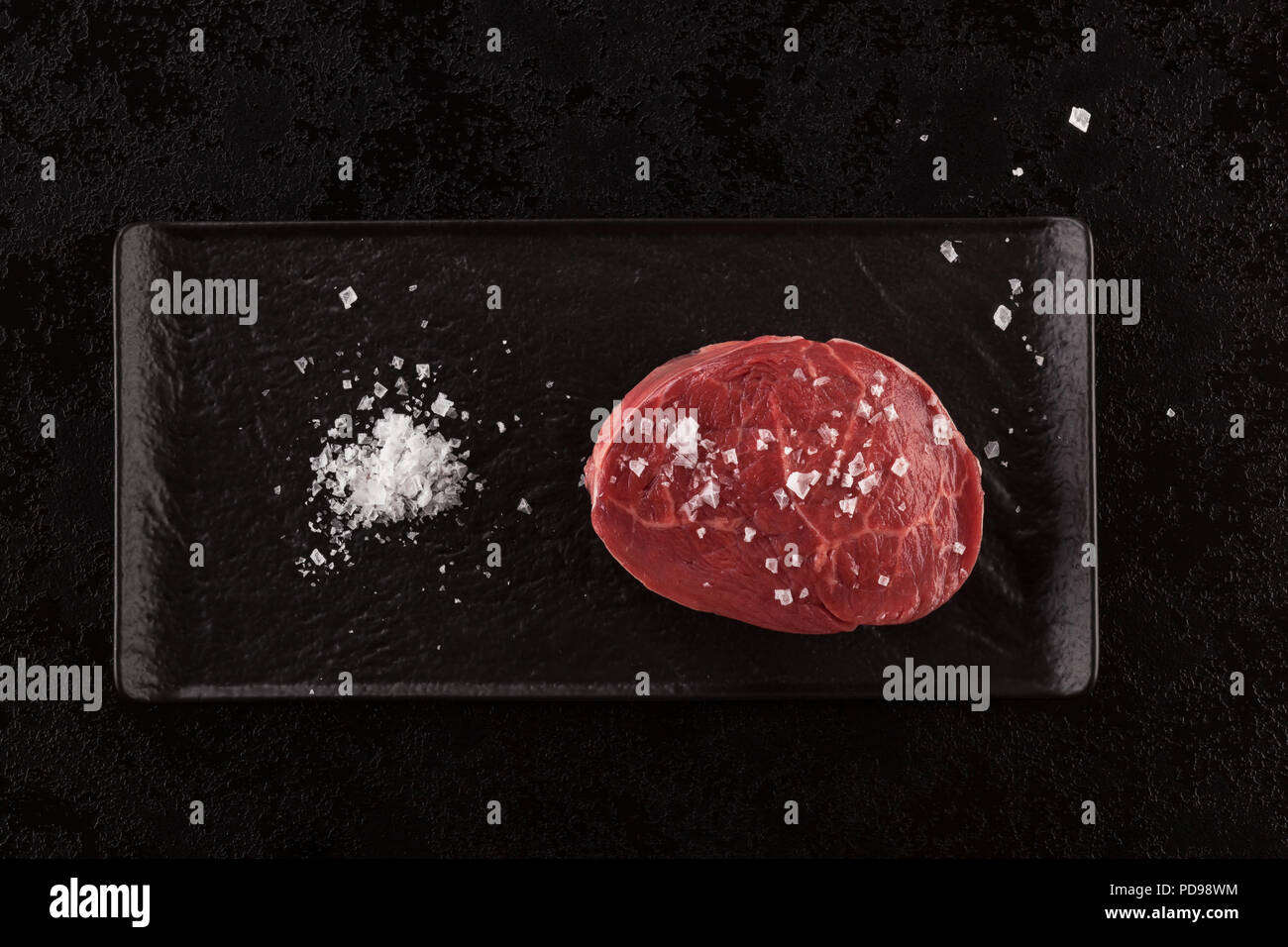 Raw beef steak with salt flakes on a black plait on dark table ...