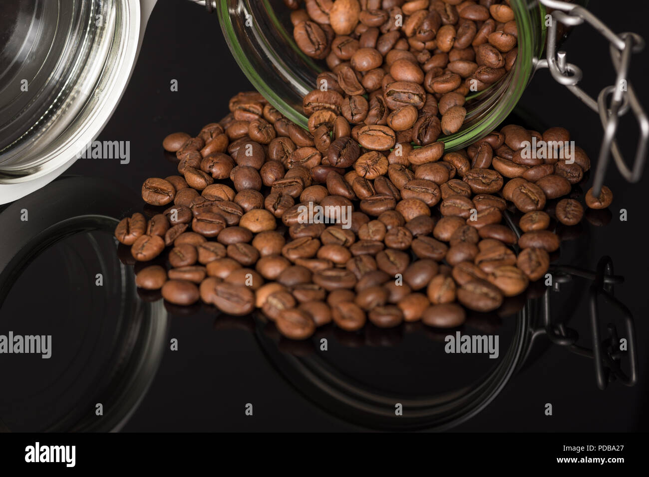 Coffee beans spilling from a glass storage jar onto black reflective ...