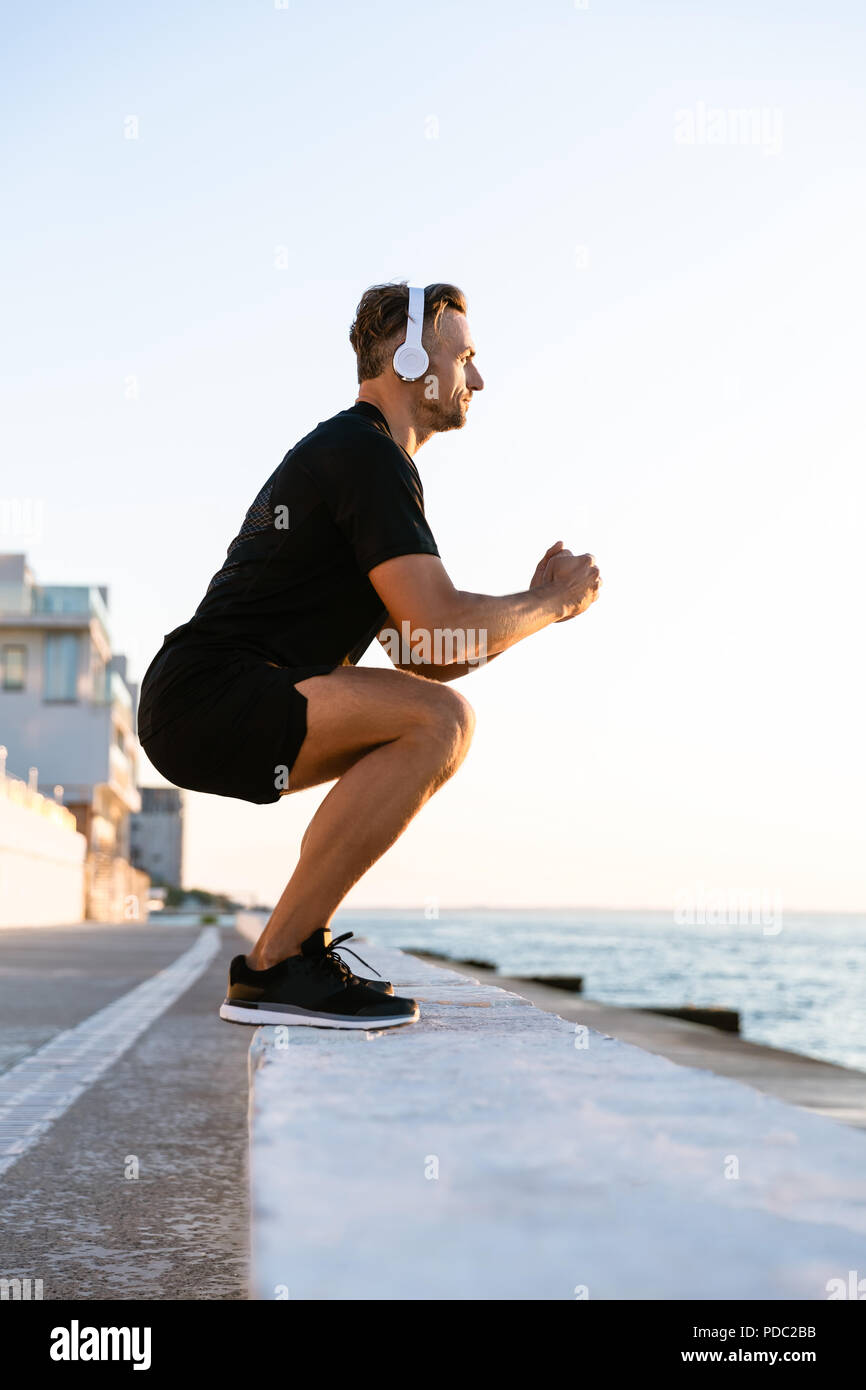 side view of athletic adult man in headphones standing squats on ...