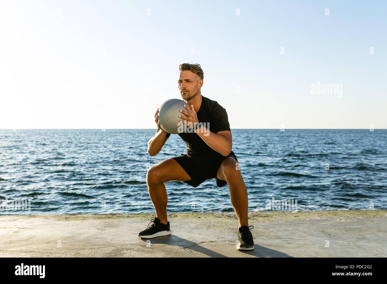 sporty adult man doing squats with fit ball on seashore Stock Photo - Alamy