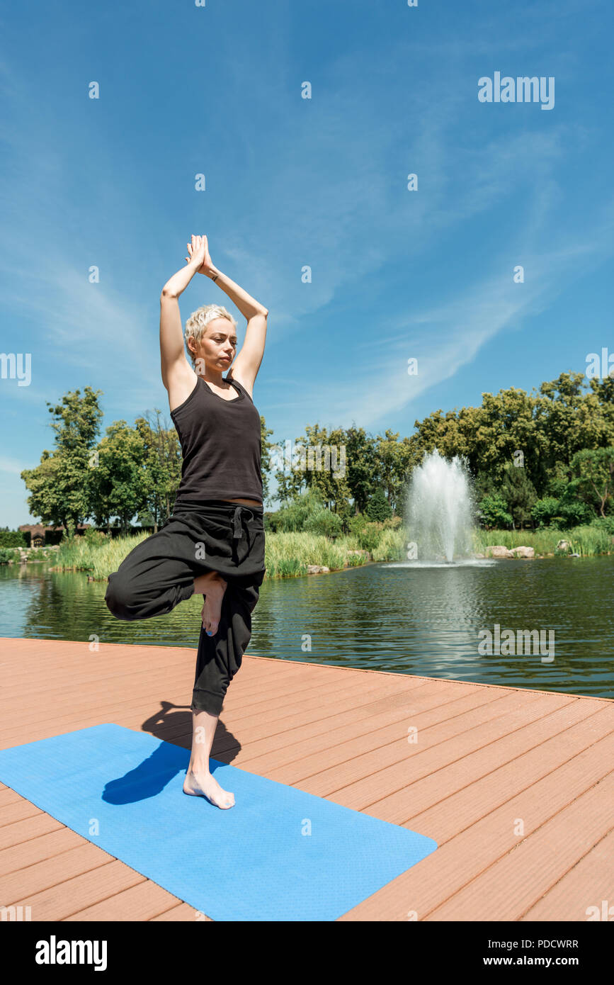 woman practicing yoga in Tree pose (Vrksasana) on yoga mat near river ...