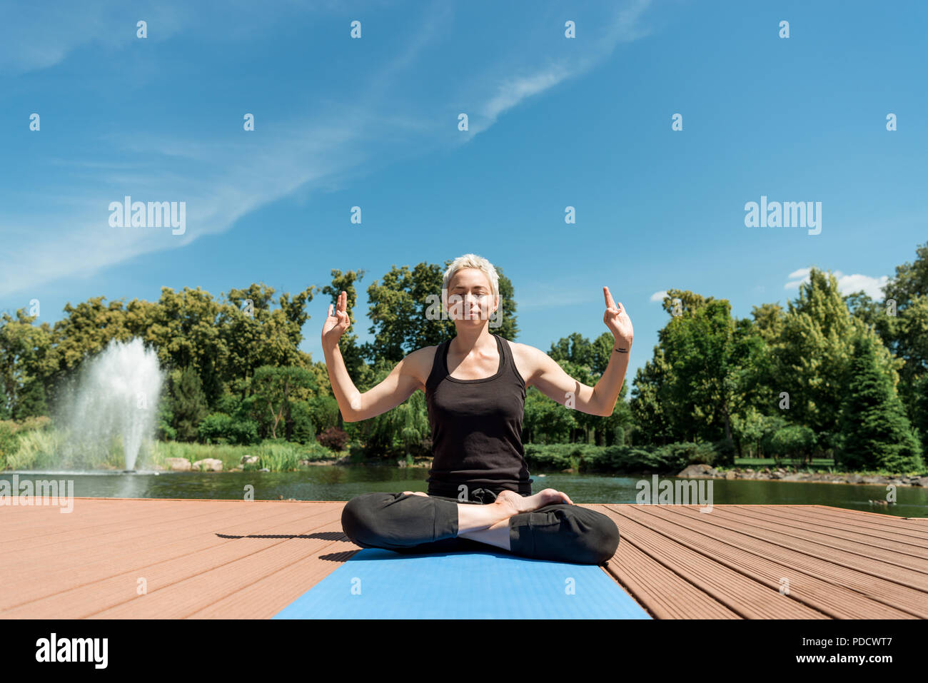 happy woman practicing yoga in lotus pose on yoga mat near river in ...
