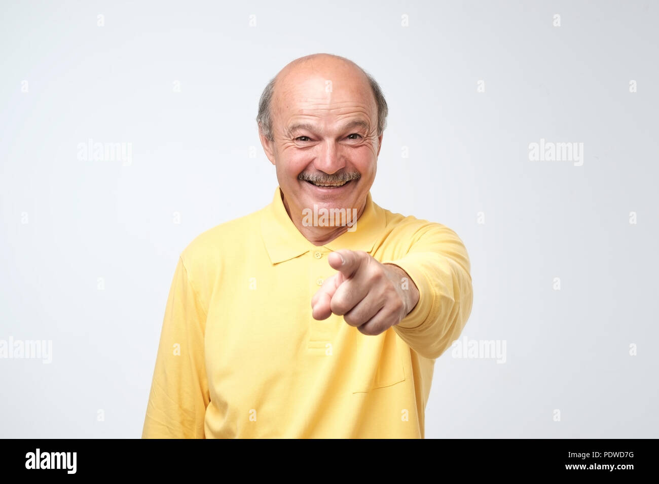 Cheerful senior man in yellow clothes indicates happily at you, chooses ...