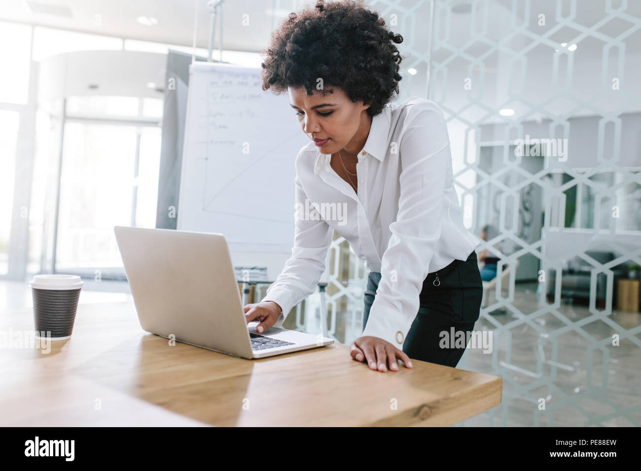 African young woman standing by table and looking at her laptop ...